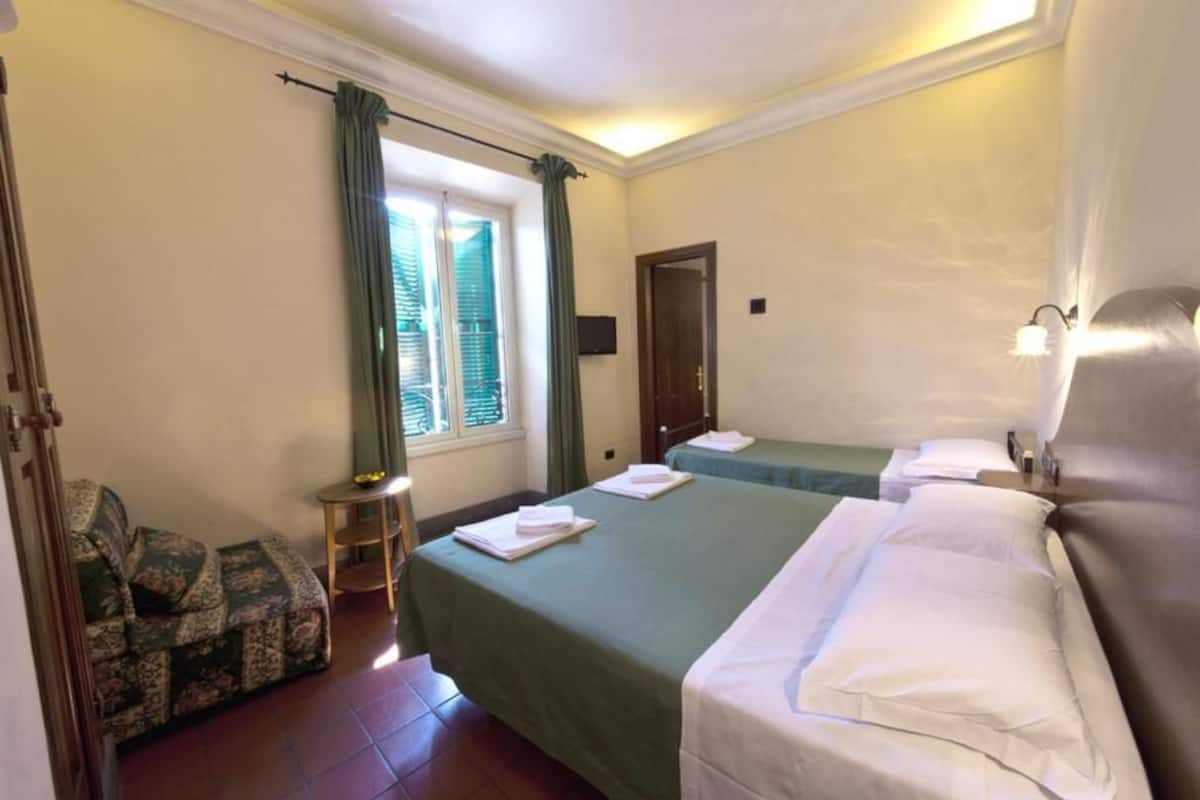 Quarto de hotel com duas camas, uma mesa pequena, uma cadeira, janela com cortinas e luminária na parede.