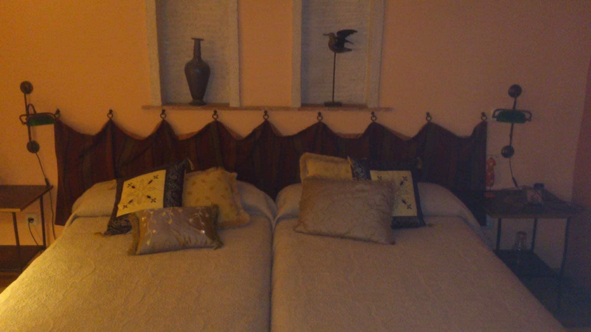 Uma cama de casal com duas mesas de cabeceira, cada uma com uma luminária, em quarto com cabeceira de madeira e janela com um vaso e uma escultura de pássaro.