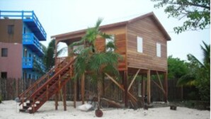 Uma casa de madeira com uma escada numa praia de areia, rodeada por palmeiras e um edifício azul ao fundo.