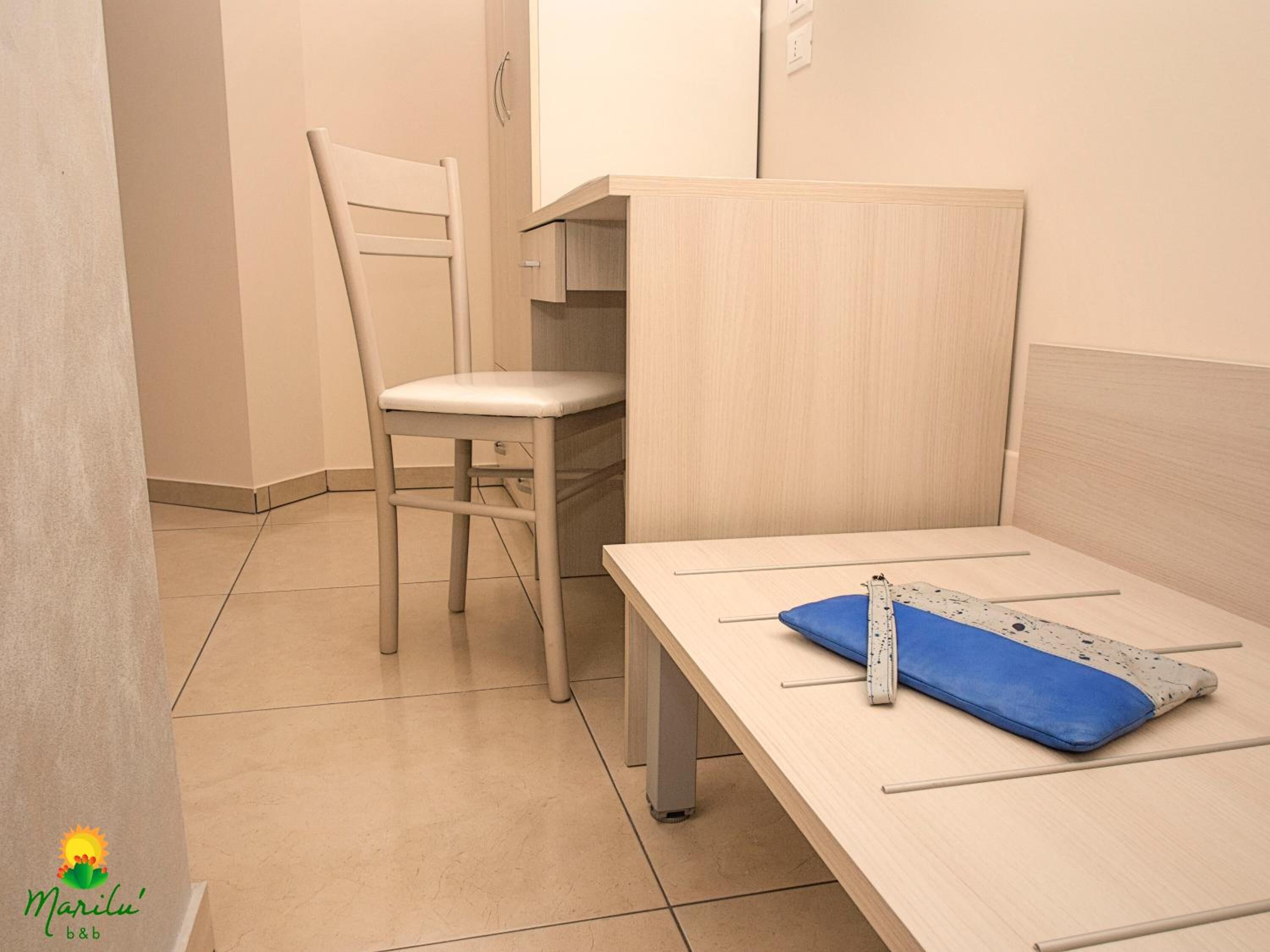 Um quarto pequeno e bem organizado, com uma cadeira, uma mesa com almofada azul e um armário de madeira.