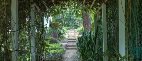 Uma trilha de pedra serpenteia por um jardim exuberante, com uma galeria coberta acima.