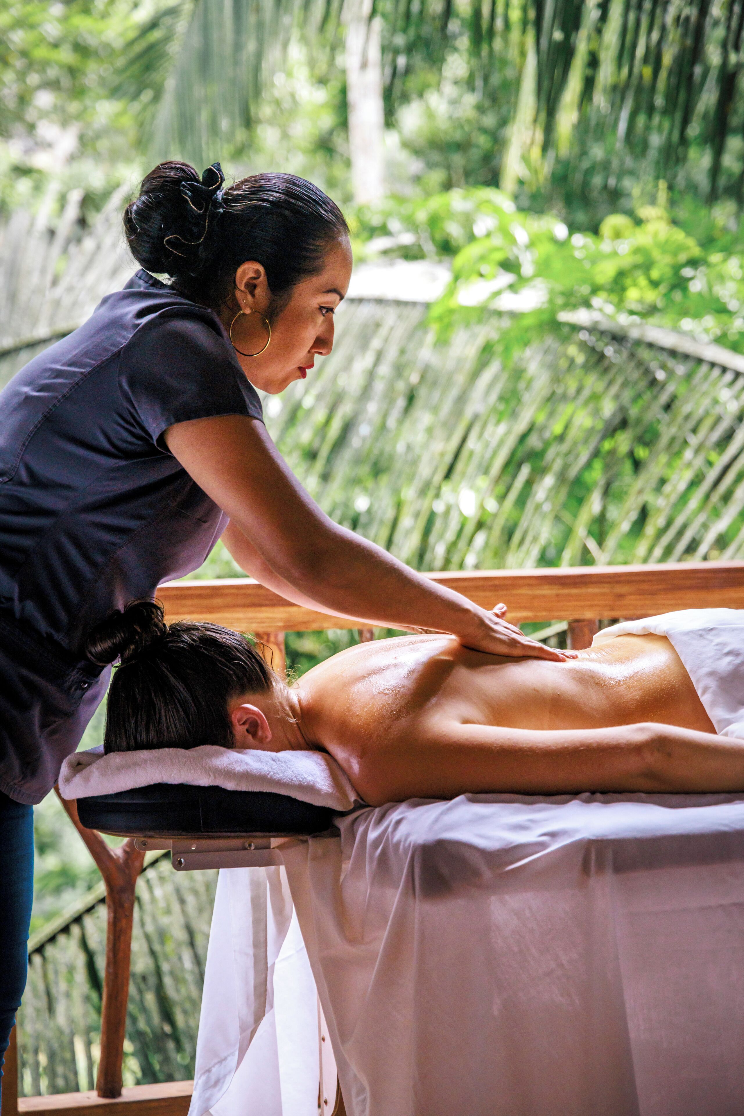 Uma pessoa recebendo massagem em um quarto com cama de madeira e vista para vegetação exuberante.