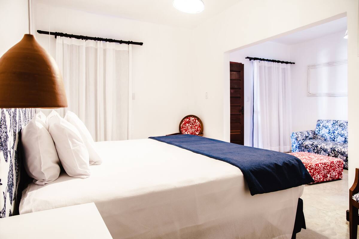 Quarto de hotel com cama, luminária pendente, cadeira e porta que dá para outro cômodo.
