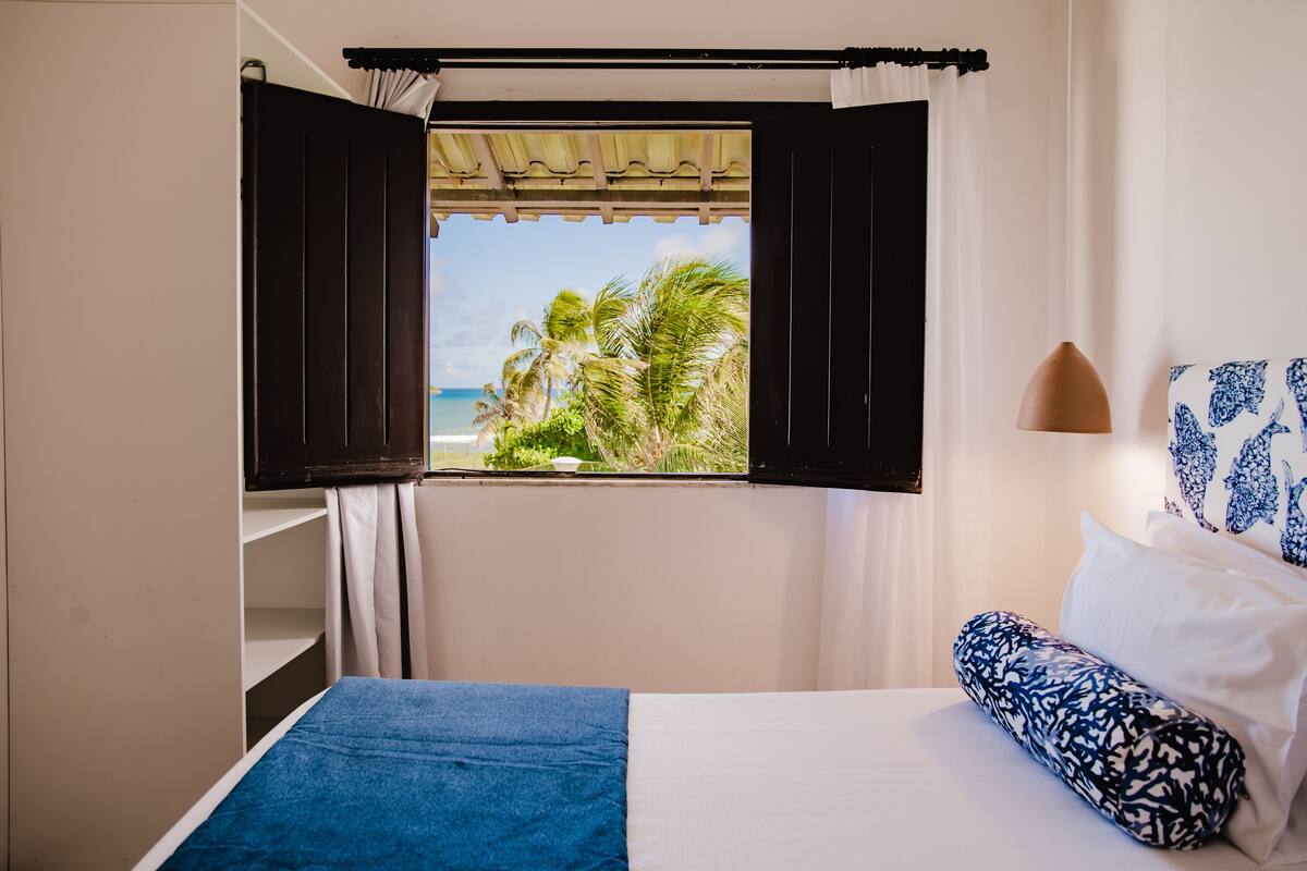 Quarto de hotel com cama, janela com persianas abertas, vista para palmeiras e oceano, e almofada estampada.