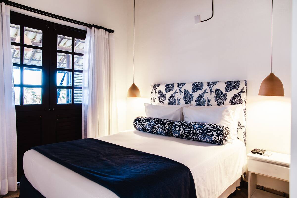 Cama bem arrumada com cabeceira estampada, mesa de cabeceira com abajur e janela com cortinas.