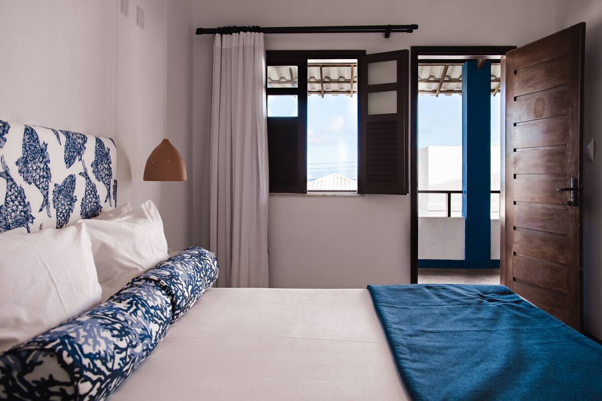 Quarto com cama, almofadas estampadas em branco e azul, manta azul, luminária pendente e vista para o exterior por uma porta aberta.