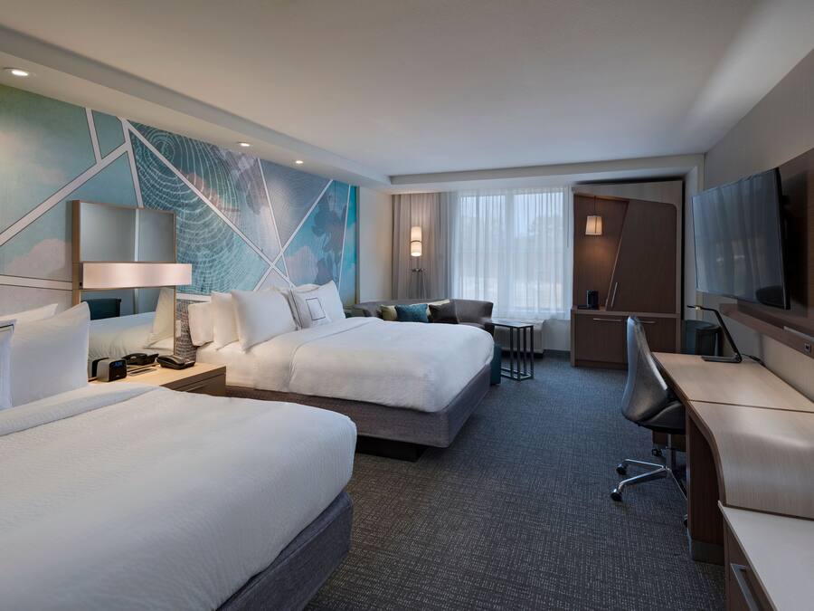 Quarto de hotel com duas camas, uma escrivaninha, uma cadeira, uma televisão e uma janela grande.