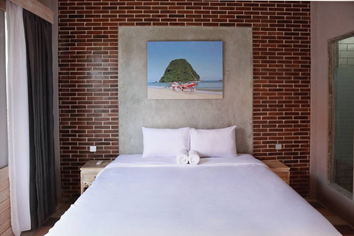 Quarto de hotel com parede de tijolos, cama com roupa de cama branca, pintura de uma praia e janela com cortinas.