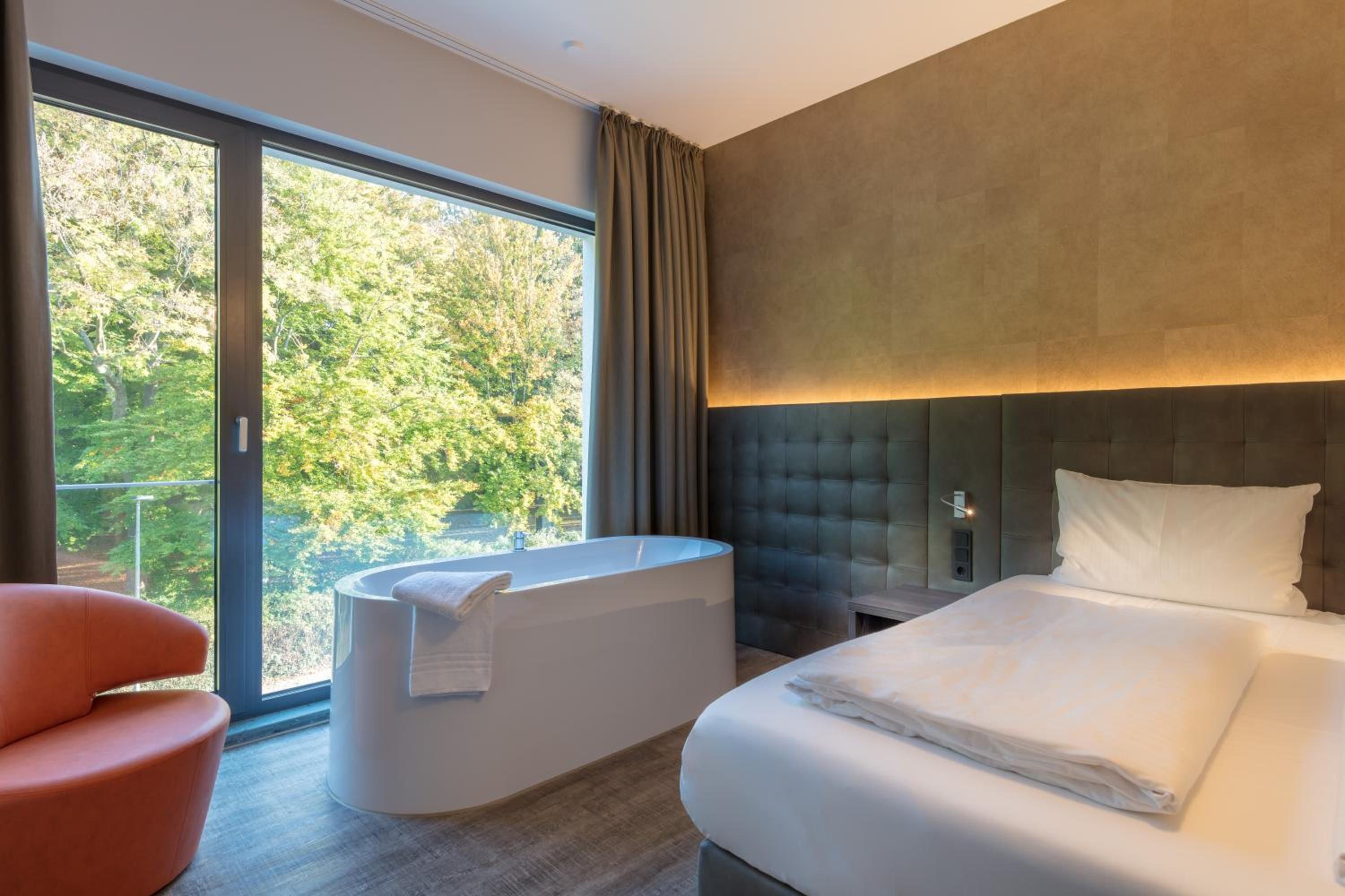 Quarto de hotel moderno com uma grande janela, uma banheira, uma cama e um espaço de estar.