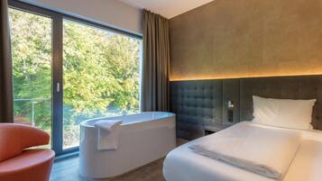 Quarto de hotel moderno com uma grande janela, uma banheira, uma cama e um espaço de estar.