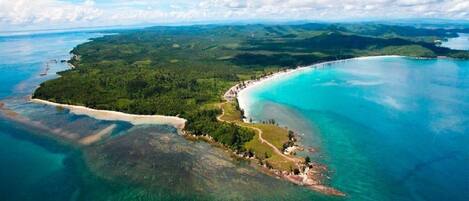 Vista aérea de uma área costeira com praia, águas turquesa cristalinas e vegetação verde exuberante.