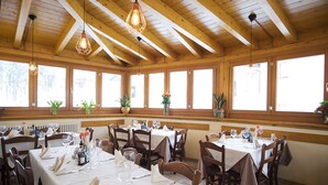 Um restaurante com vigas de madeira e grandes janelas, mesas dispostas com toalhas brancas e copos de vinho.