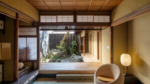 Um quarto japonês tradicional com tatames, uma mesa de madeira baixa, uma cadeira de fibra natural e vista para um jardim através de uma porta shoji.