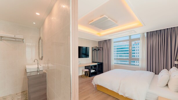 Quarto de hotel moderno com uma cama grande, uma escrivaninha com televisão de ecrã plano, uma casa de banho com lavatório e espelho, e vista da cidade através de grandes janelas.