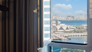 Vista da janela sobre um horizonte urbano com edifícios altos, uma orla marítima e uma estrutura arquitetônica marcante.