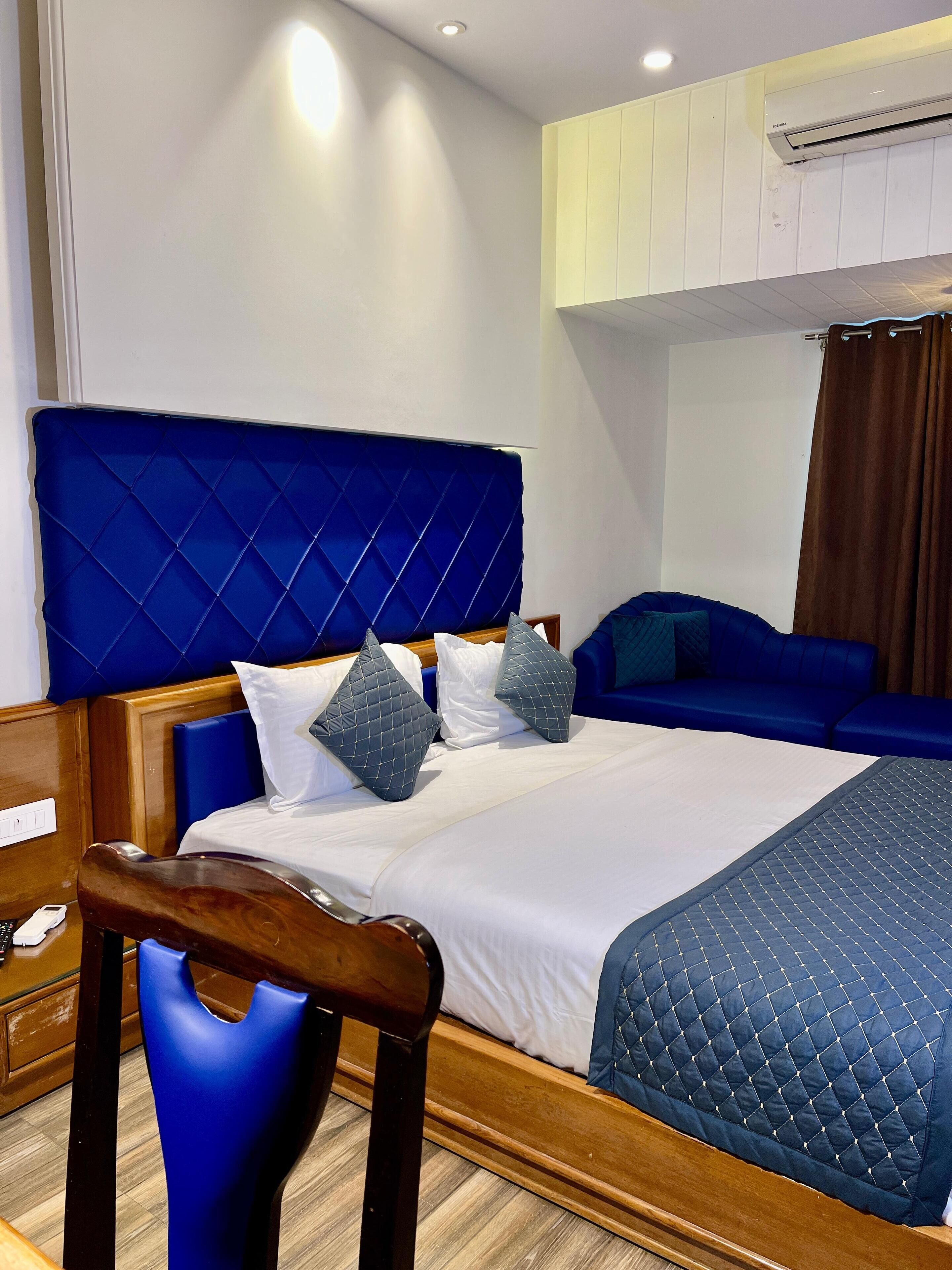 Quarto de hotel com uma cama grande, cabeceira de madeira, sofá azul estofado e ar condicionado branco na parede.