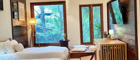 Quarto com duas camas, uma escrivaninha de madeira, tapete tecido e grandes janelas com vista para o verde.