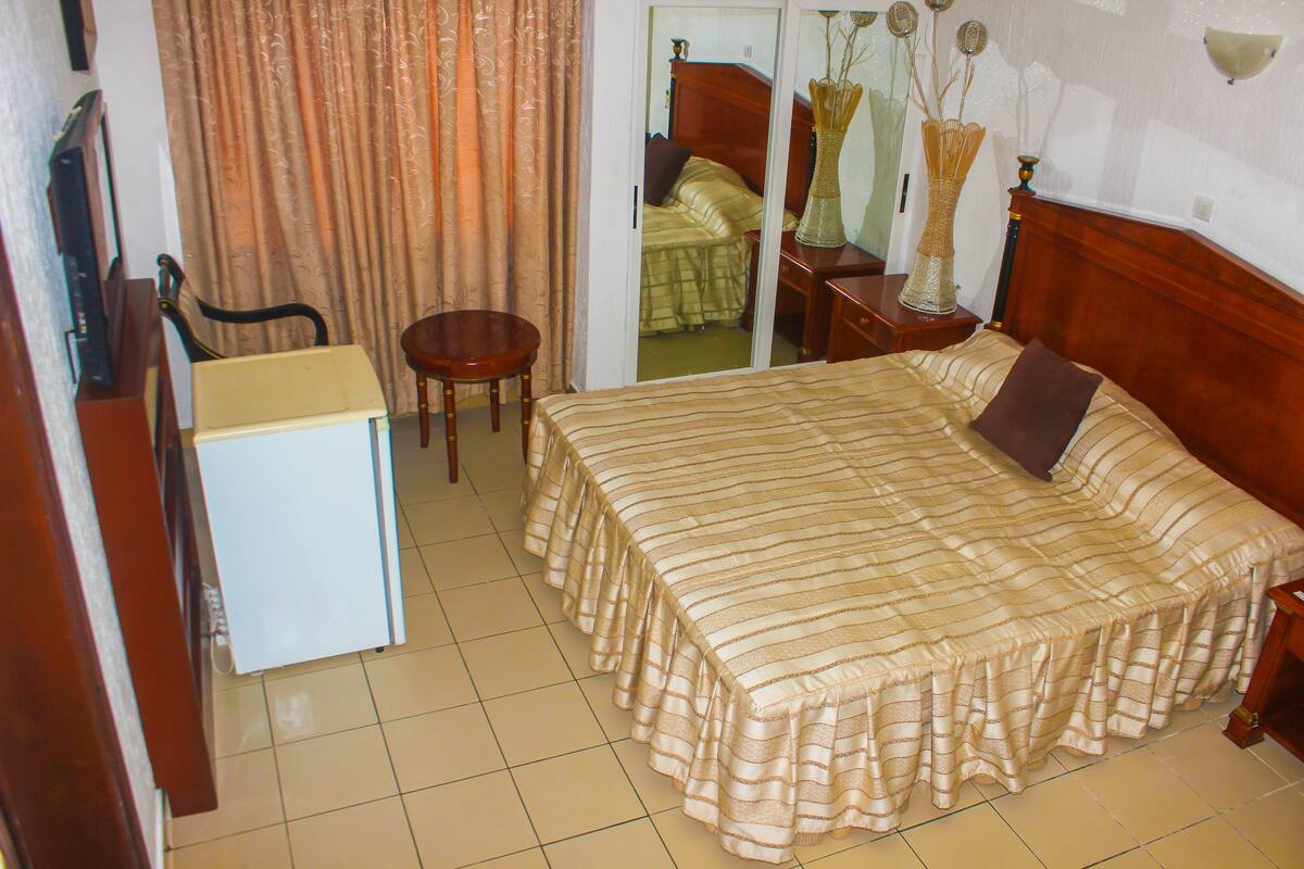 Quarto de hotel com cama, uma pequena mesa de trabalho com cadeira, frigorífico e espelho.