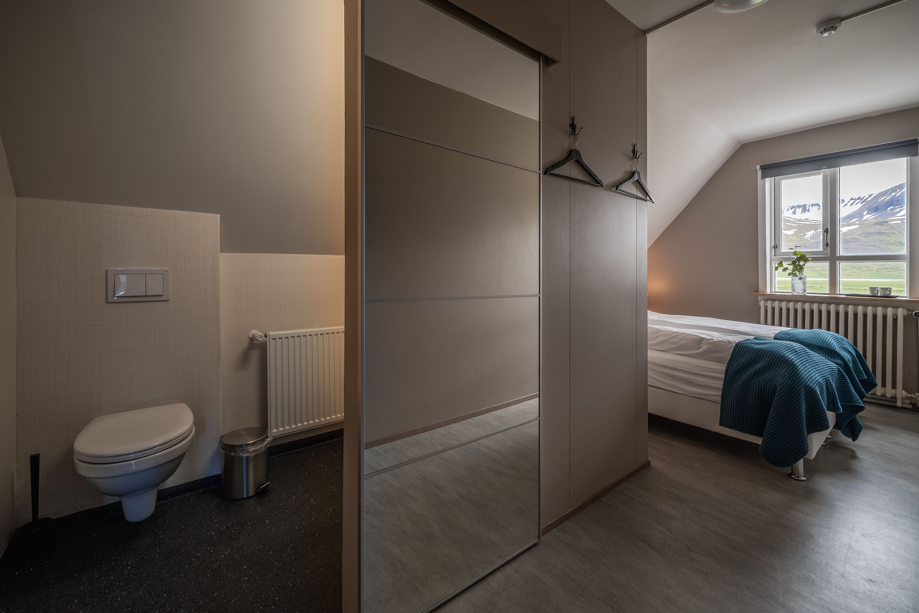 Quarto moderno com cama, janela com vista para montanhas e banheiro com vaso sanitário e radiador.
