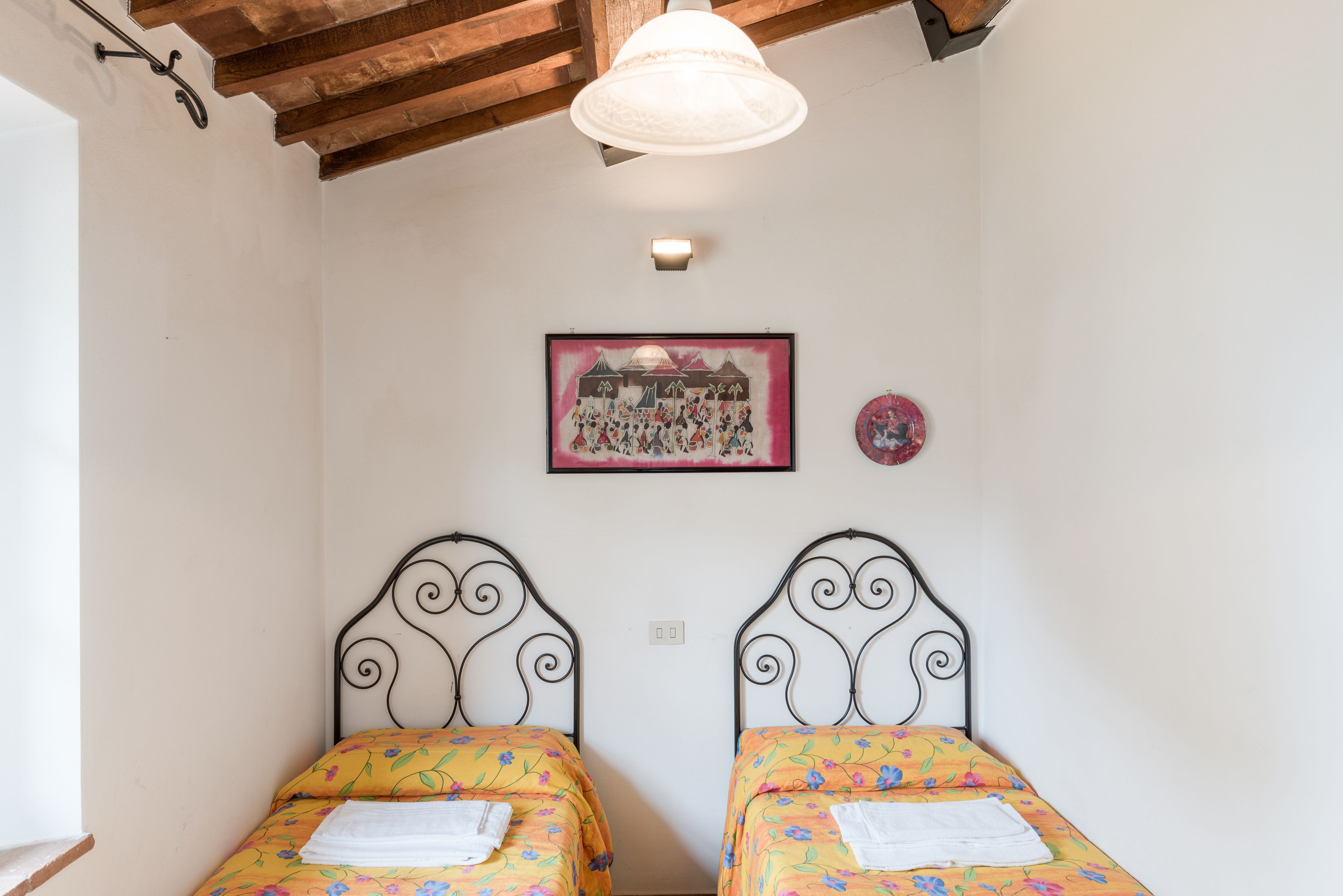 Quarto com duas camas, vigas de madeira no teto, uma luminária fixada na parede e um quadro emoldurado.