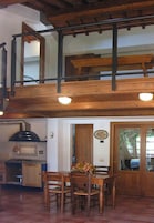 Uma sala de jantar com mesa e cadeiras de madeira, uma cozinha com fogão e armários, e uma escada que leva a um andar superior.