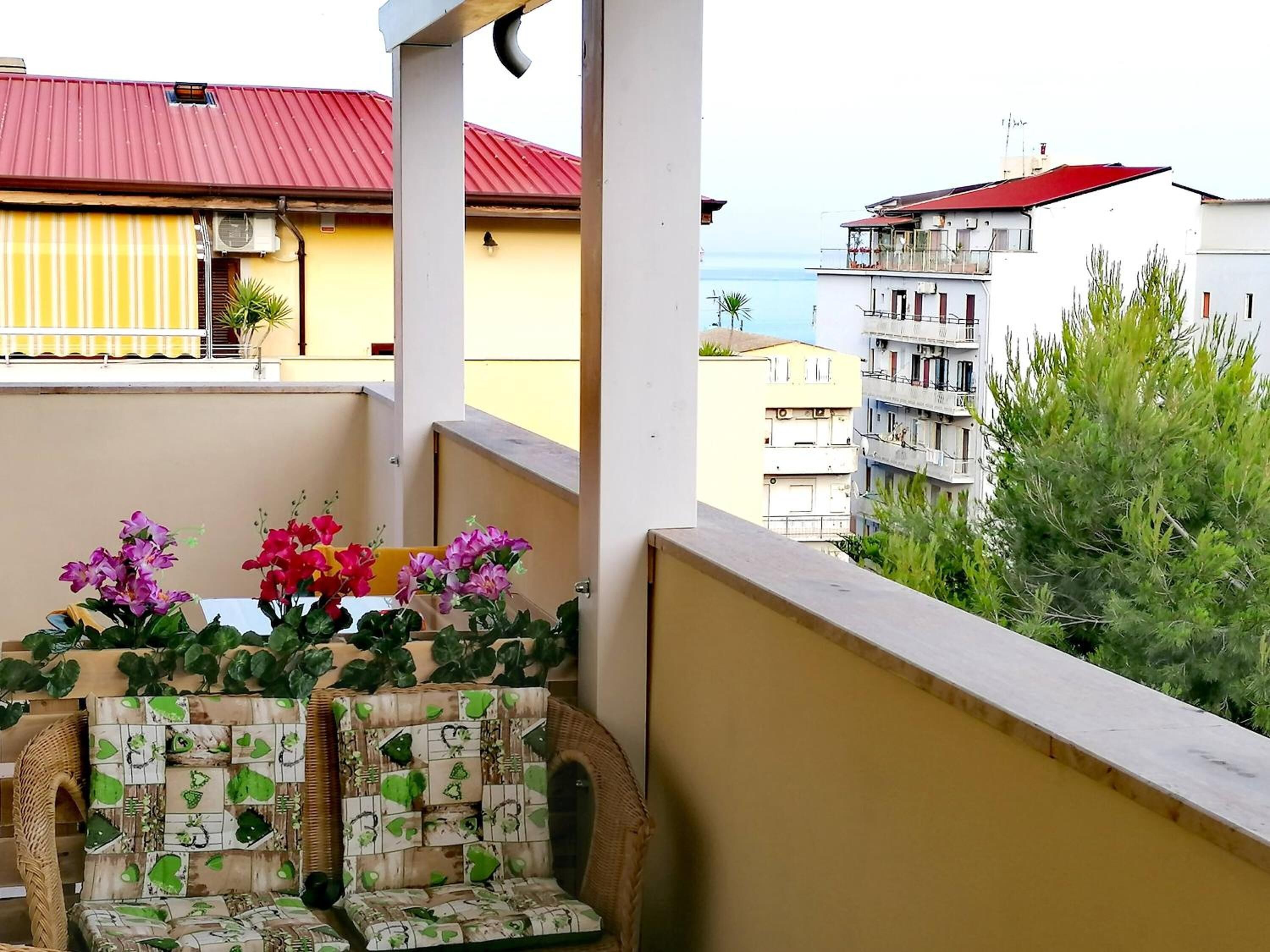 Varanda com mobiliário de vime e plantas em vasos, com vista para uma área residencial, edifícios e o mar.