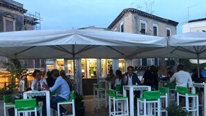 Um café ao ar livre com mesas brancas e cadeiras verdes, cercado por guarda-sóis.