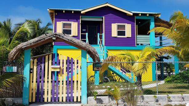 Uma casa colorida com fachada roxa e amarela, um portão de madeira e um telhado de palha.