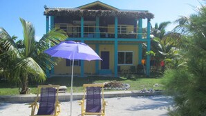 Um edifício de dois andares com telhado de palha, localizado na praia, e duas cadeiras de praia com um guarda-sol roxo.