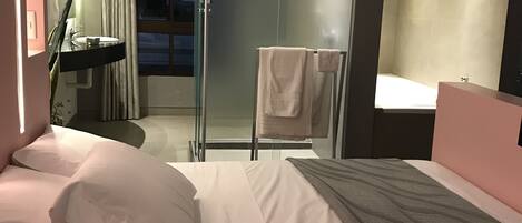 Quarto de hotel moderno com uma cama grande, box de vidro, toucador com espelho e vista para a paisagem urbana.