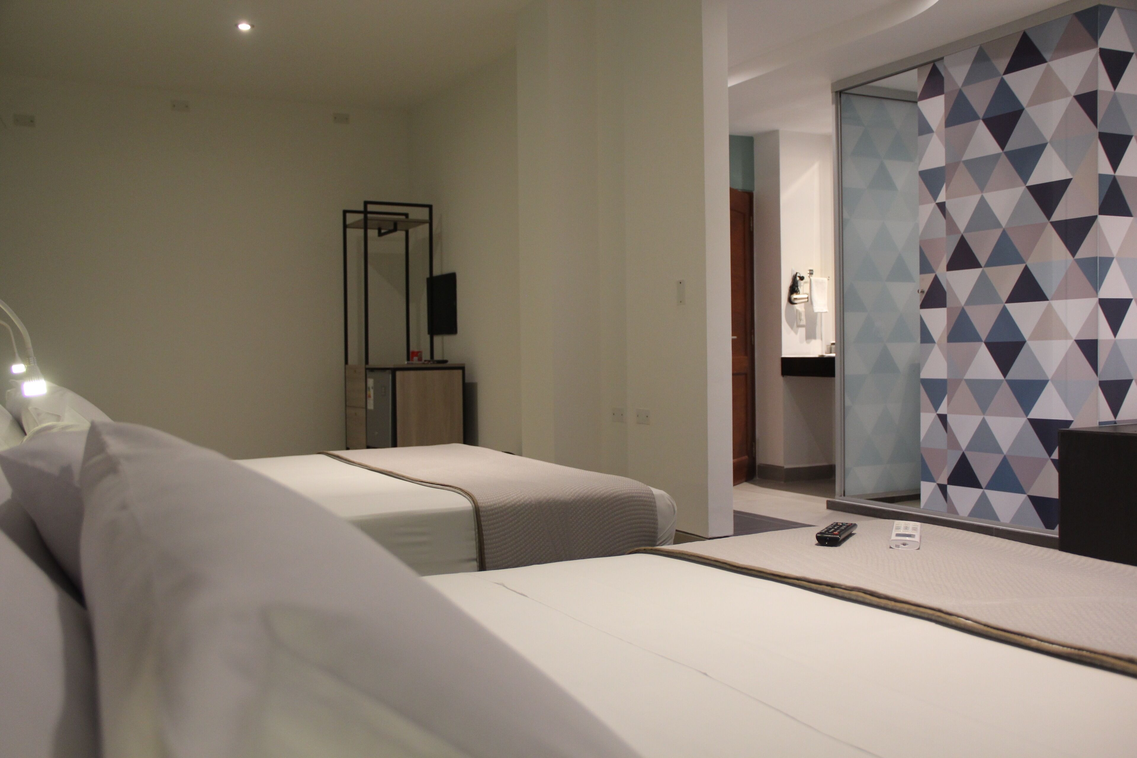 Quarto de hotel com duas camas, um espelho, uma mesinha e um banheiro visível através de uma porta de vidro.