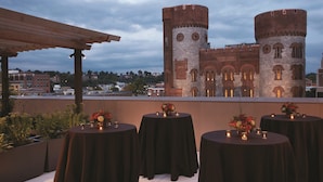 Terraço no telhado com mesas redondas preparadas para refeições, com vista para um edifício histórico com duas torres imponentes.