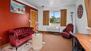Quarto com sofá vermelho, mesa de madeira, poltrona vermelha, mesinha redonda com planta, alvo de dardos e pintura de uma paisagem costeira.