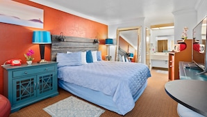Quarto de hotel com cama, mesa de cabeceira, escrivaninha, espelho e banheiro visível por uma porta aberta.