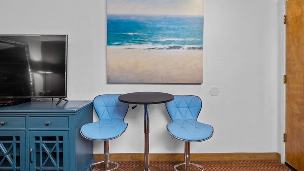 Quarto com televisão, duas cadeiras azuis, uma mesa pequena e uma pintura do oceano.