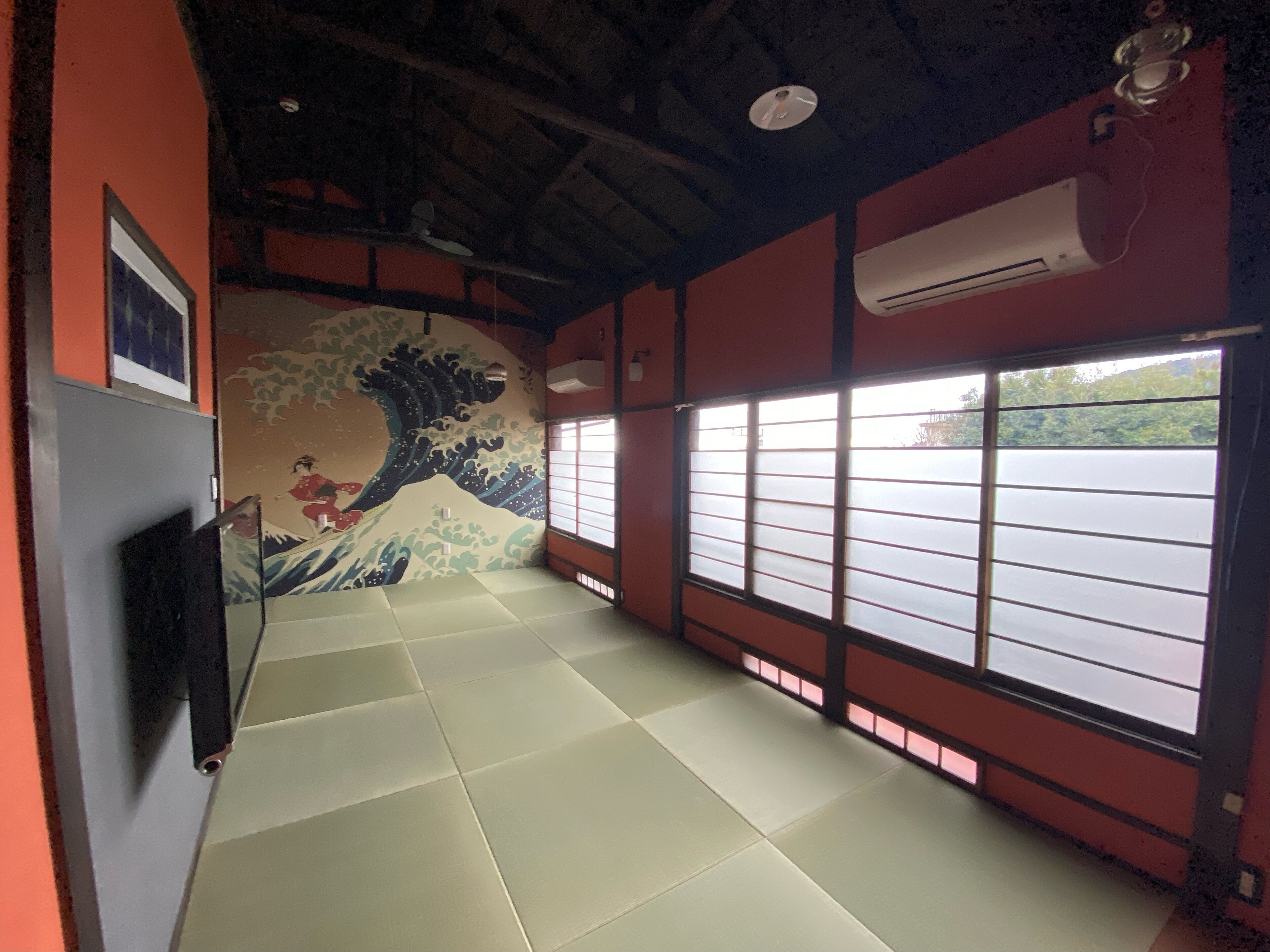 Um espaço com esteiras de tatame, um mural japonês tradicional, um ventilador de teto e uma grande janela com vista para o verde.