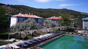 Uma área à beira da piscina com espreguiçadeiras e piscina, cercada por edifícios com telhados vermelhos e uma paisagem montanhosa ao fundo.
