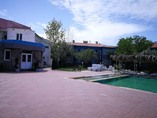 Uma área de piscina com espreguiçadeiras e uma pequena mesa, cercada por um pátio pavimentado e um edifício com telhado vermelho.