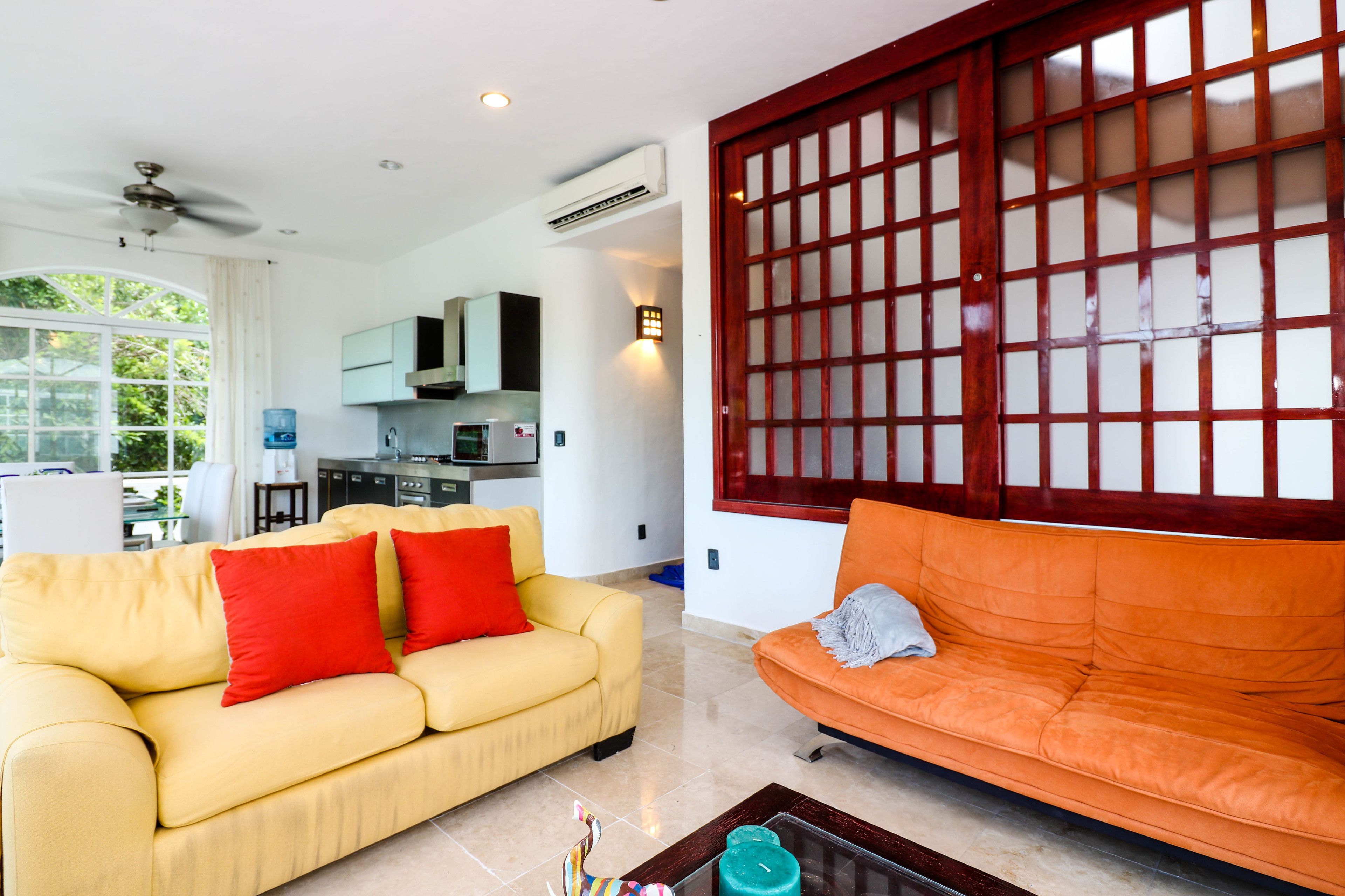 Uma sala de estar com um sofá amarelo e um sofá laranja, uma divisória de madeira e uma área de cozinha ao fundo.