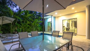 Área de piscina externa com mesa e cadeiras de vidro, cercada por palmeiras e uma casa moderna ao fundo.