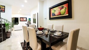 Uma sala de jantar com uma mesa de madeira escura, cadeiras brancas e um quadro com tema de frutas na parede.