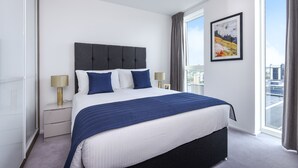 Quarto de hotel moderno com uma cama grande, mesa de cabeceira com abajur, janela com vista para a cidade e um quadro na parede.