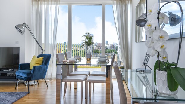 Sala de estar moderna com mesa de jantar de vidro, cadeiras azuis e amarelas, televisão e uma grande janela com vista para a cidade.