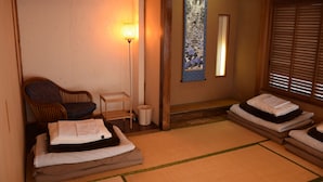 Um quarto japonês tradicional com tatames, uma cama de madeira baixa, uma cadeira, uma mesa pequena e uma obra de arte na parede.
