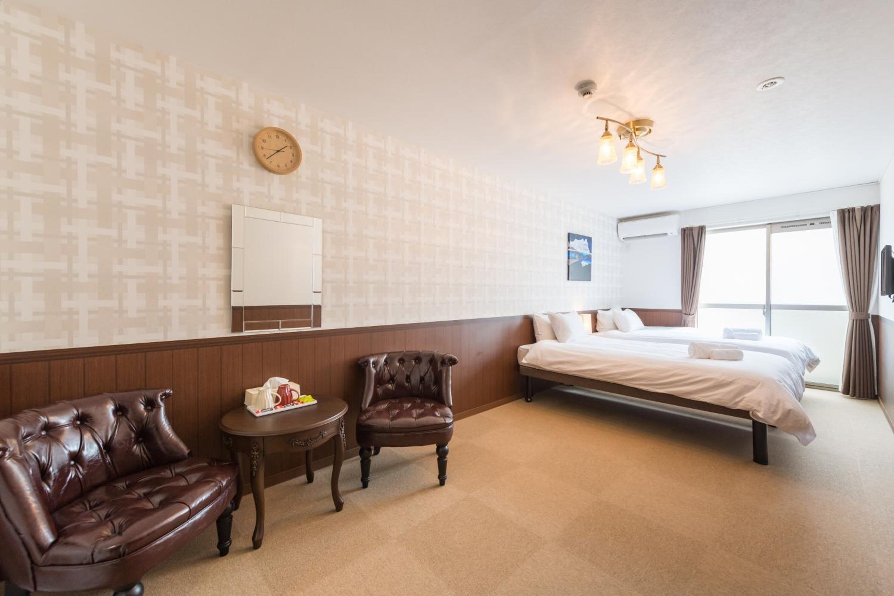 Quarto de hotel com uma cama grande, duas poltronas de couro, uma mesinha com telefone e uma chávena, um relógio na parede e uma janela com cortinas.