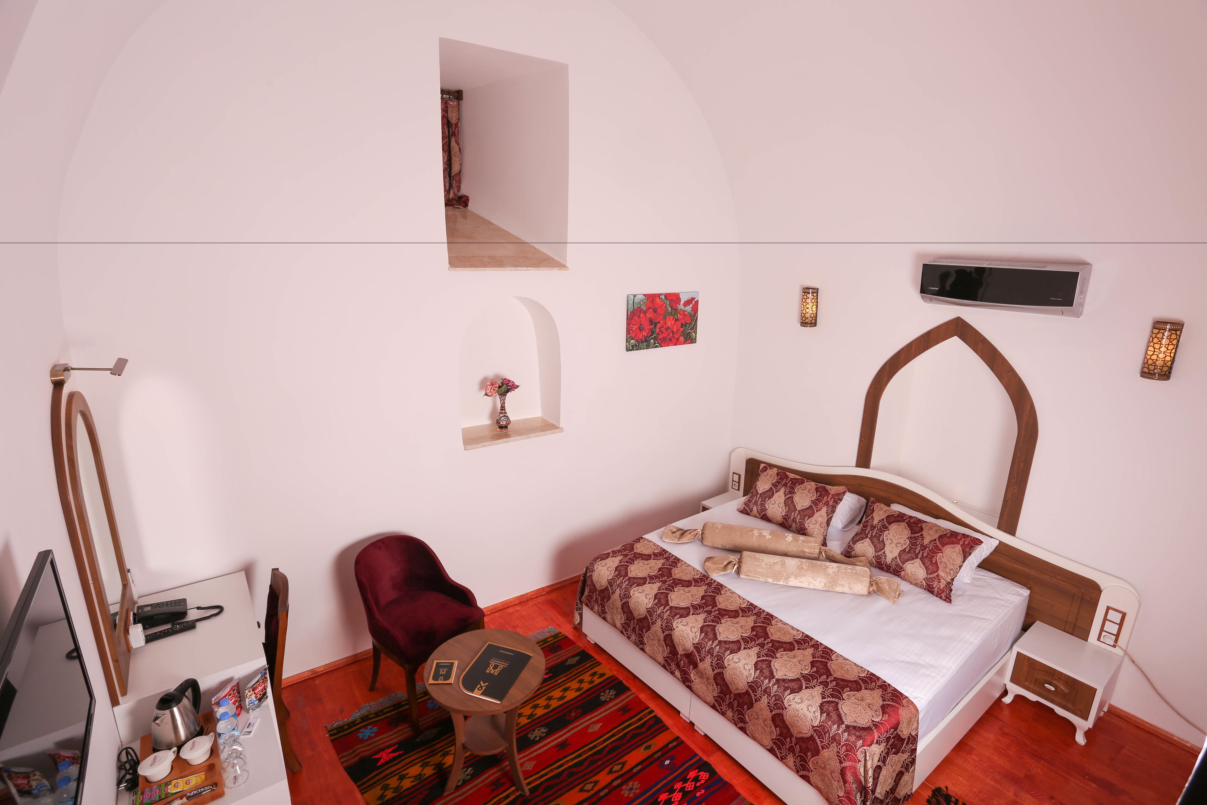 Quarto com cama, poltrona, mesinha, espelho, prateleira com um vaso e uma janela.