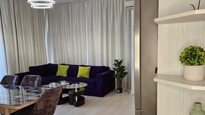 Uma sala de estar moderna com um sofá azul escuro, uma mesa de jantar de vidro e uma cozinha minimalista.