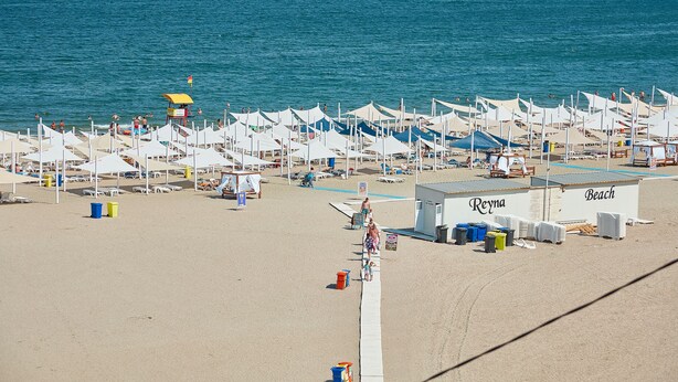 Uma praia com fileiras de guarda-sóis e cadeiras de praia, uma torre salva-vidas e uma placa com a inscrição 'Reyna Beach'.