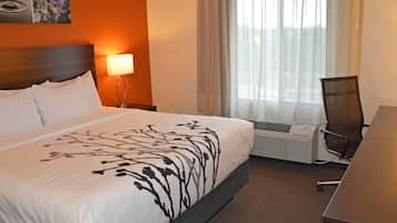 Quarto de hotel com uma cama grande, uma escrivaninha, uma cadeira, uma luminária e uma janela com cortinas.