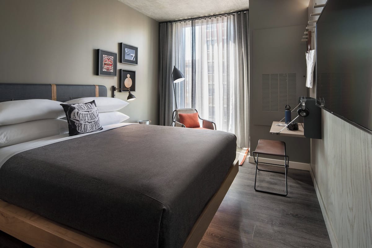 Quarto de hotel moderno com uma cama grande, uma escrivaninha com cadeira, uma televisão de tela plana e uma janela com cortinas.